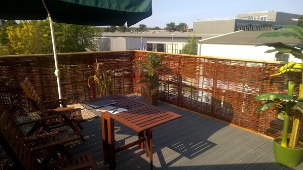 Um deck de madeira com mesa e cadeiras, cercado por uma cerca de bambu, e vista para edifícios ao fundo.