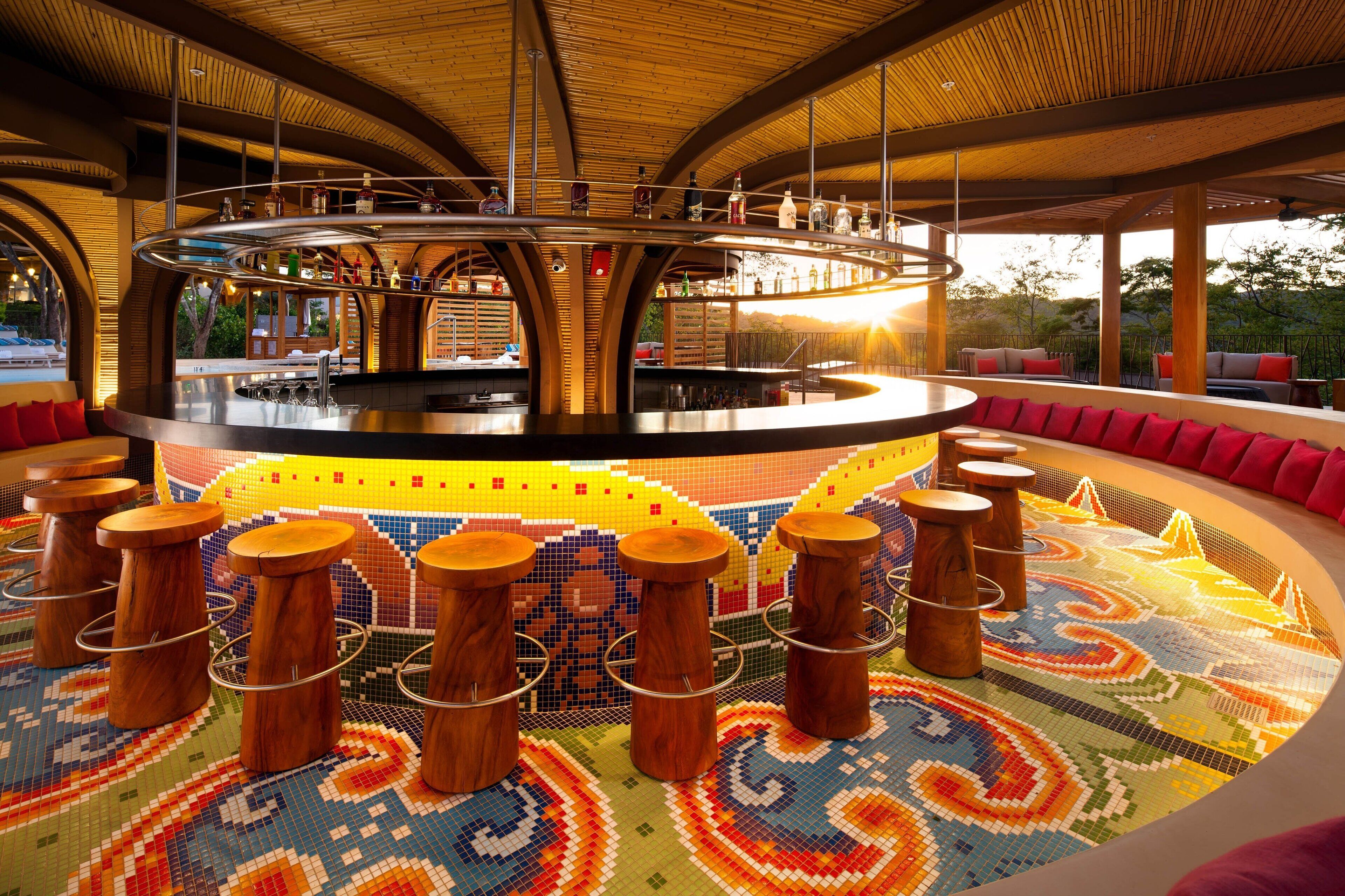 Uma área de bar com balcão circular, banquetas de madeira e um chão com mosaico colorido.