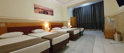 Quarto de hotel com quatro camas dispostas em fila, um quadro na parede, uma janela grande com cortinas e uma televisão fixada na parede.