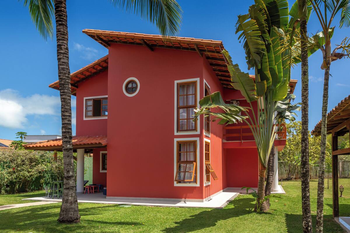 Uma casa vermelha de dois andares com telhado de telhas, rodeada por um gramado bem cuidado e palmeiras.