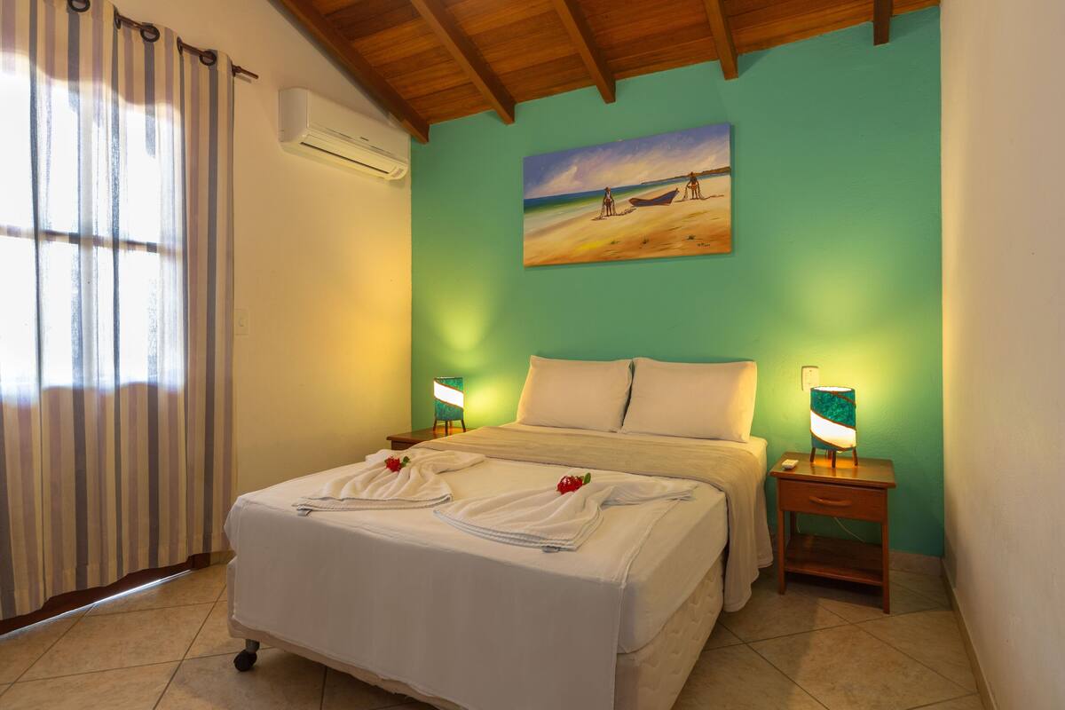 Quarto de hotel com cama, pintura de uma paisagem praiana e mesa de cabeceira com abajur.