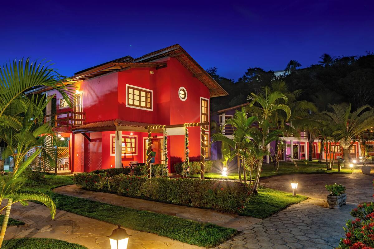 Um edifício vermelho com varanda, rodeado por jardins bem cuidados e palmeiras.