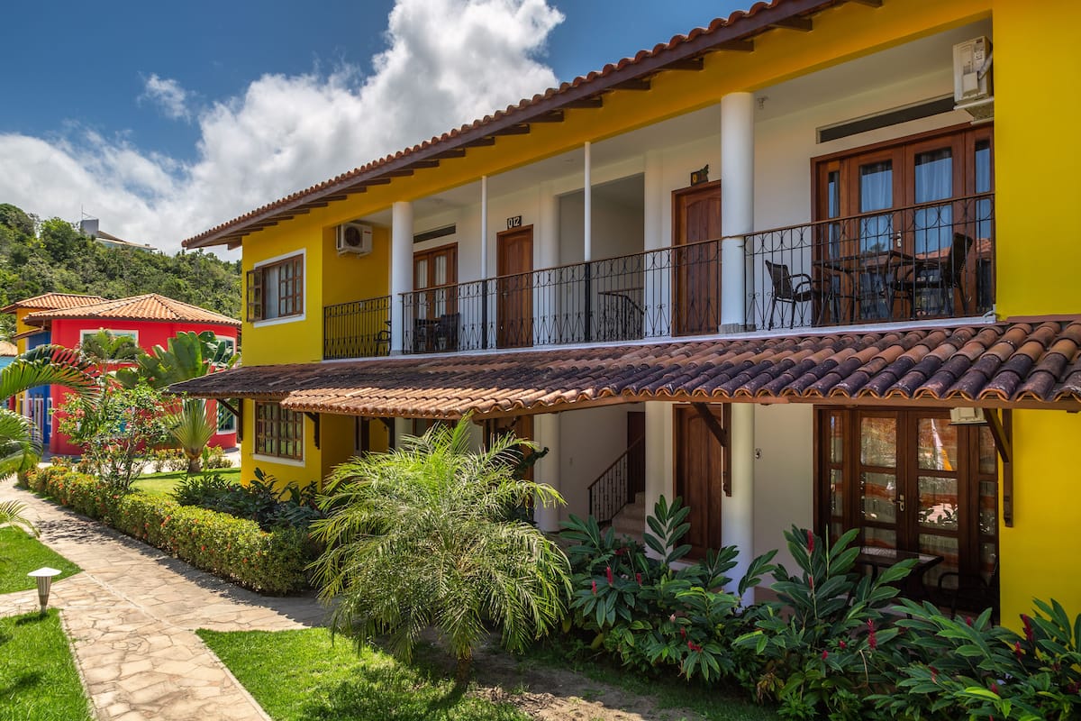 Um edifício amarelo com telhado em telhas, varanda com grades de metal e um caminho pavimentado que leva à entrada.