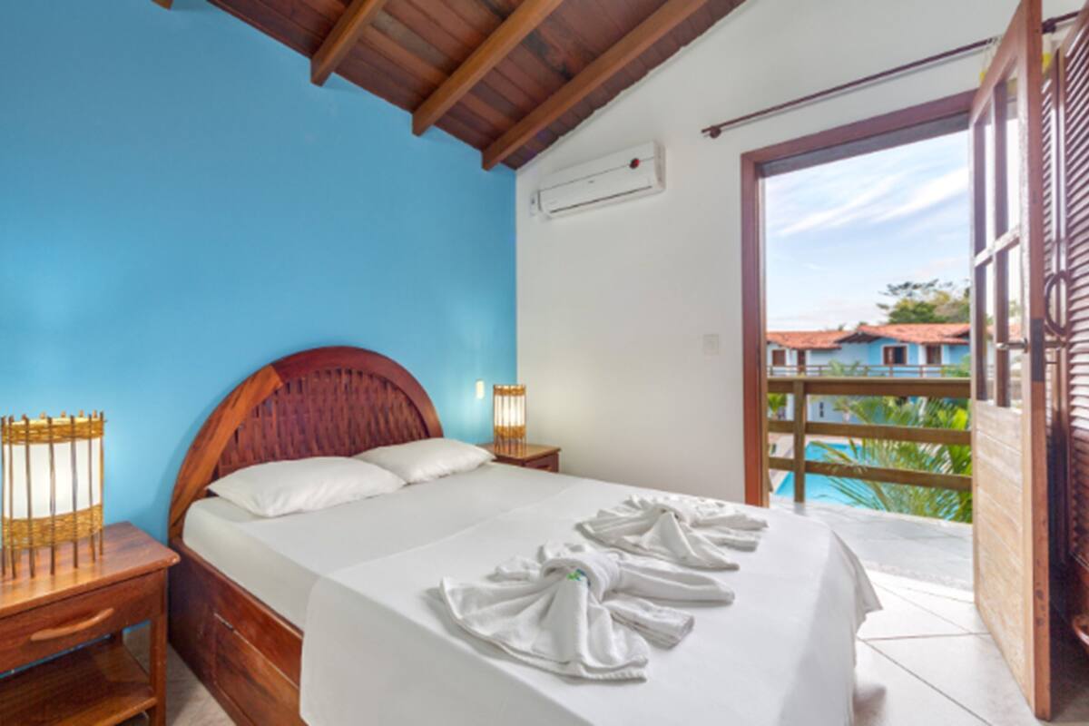 Quarto com cabeceira de madeira, cama com roupa de cama branca, mesa de cabeceira com abajur e vista para uma piscina através de uma porta aberta.