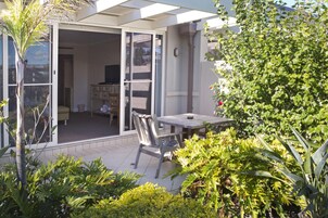 Um pátio com mesa e cadeiras, rodeado por vegetação exuberante e uma porta de vidro deslizante que dá para um espaço interior com televisão e área de estar.