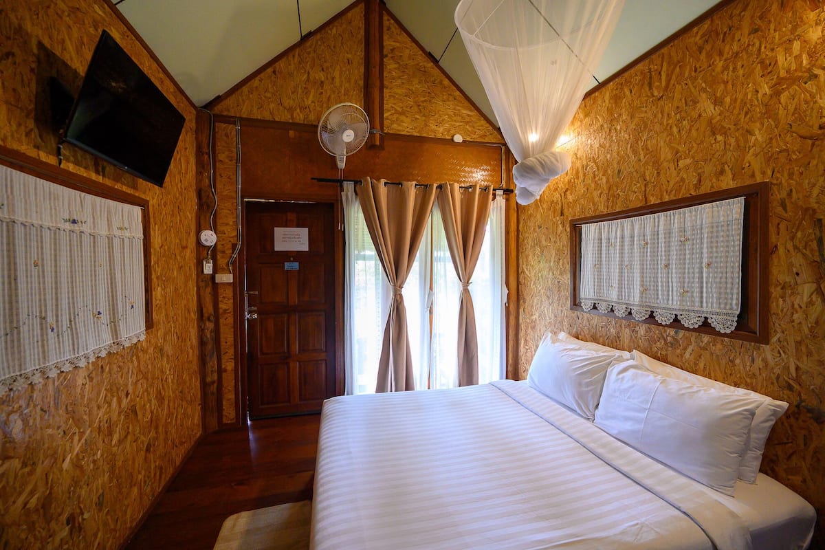 Quarto com cama, ventilador de teto, janela com cortinas e televisão presa à parede.