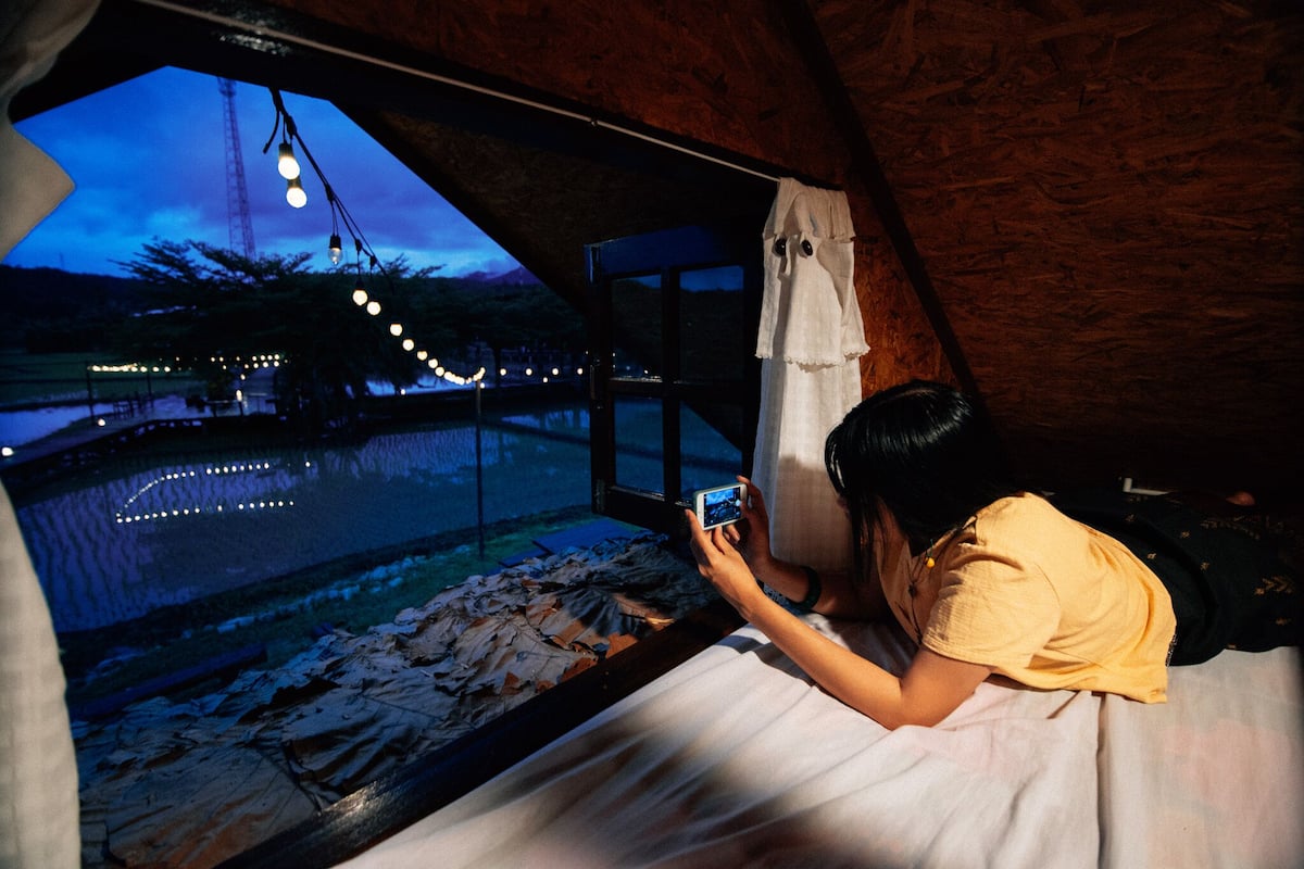 Uma pessoa deitada em uma cama em um quarto parecido com uma tenda, olhando para um corpo d'água com luzes ao longe.