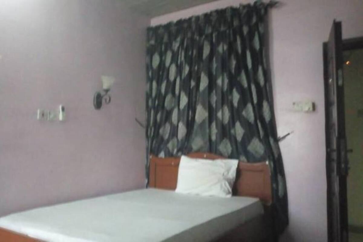 Uma cama de solteiro com colchão branco e cabeceira marrom em um quarto com paredes roxas, cortina estampada e duas luminárias fixadas na parede.