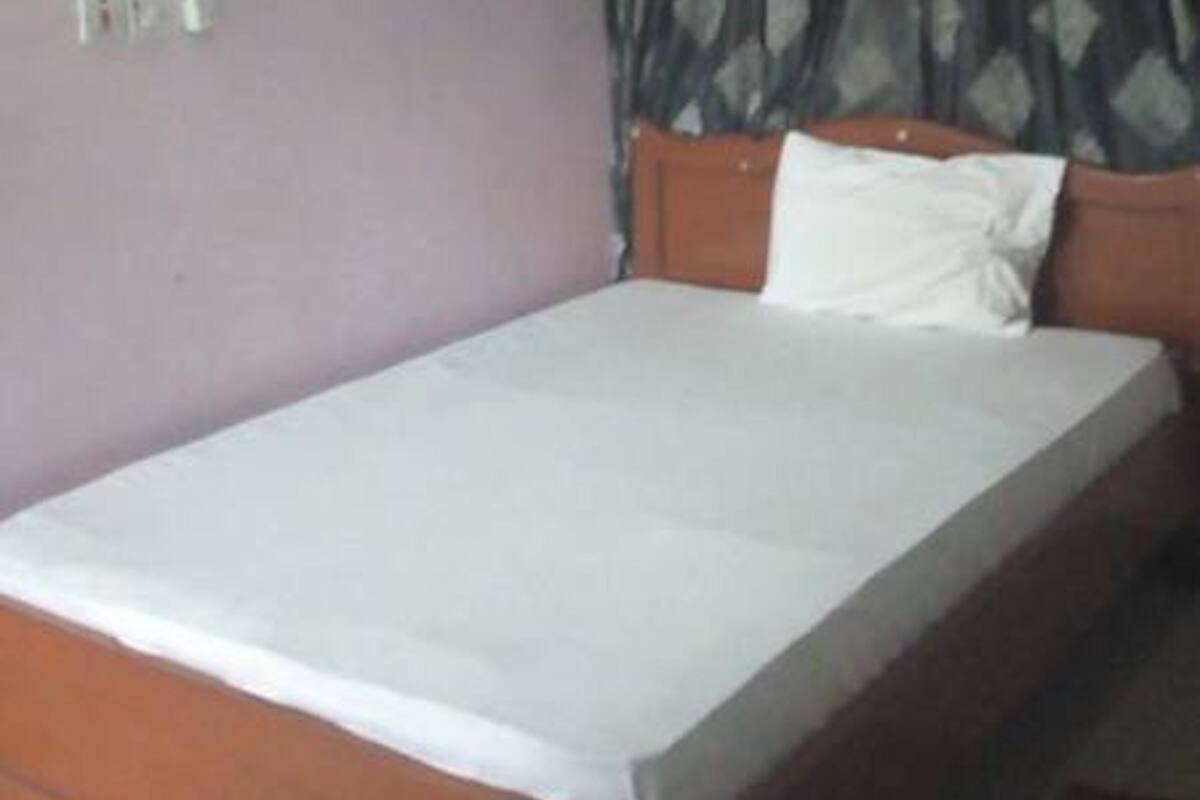Uma cama de solteiro com estrutura de madeira e roupa de cama branca em um quarto com papel de parede estampado e chão ladrilhado.