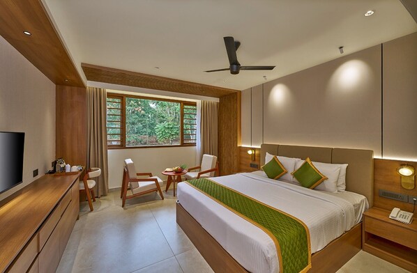Quarto de hotel com cama, escrivaninha, cadeiras, televisão e janela com vista para o verde.