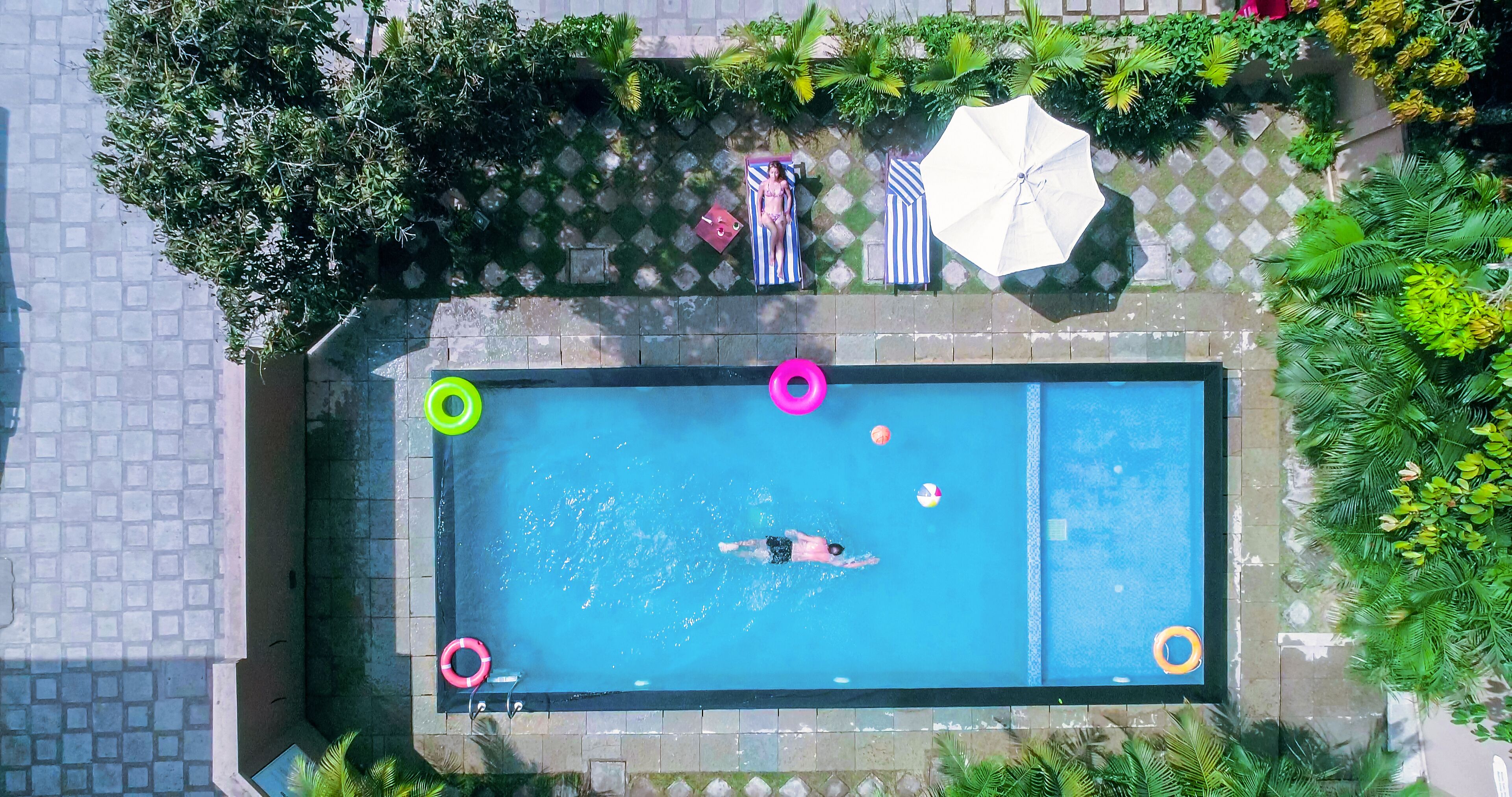 Vista aérea de uma piscina com uma pessoa nadando, cercada por vegetação e um caminho de azulejos.