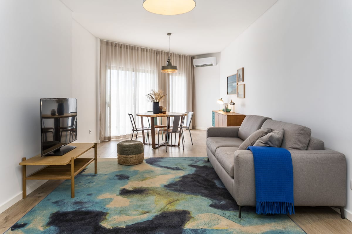Uma sala de estar moderna com um sofá, uma televisão sobre um suporte de madeira, uma mesa de jantar com cadeiras e uma janela grande com cortinas.
