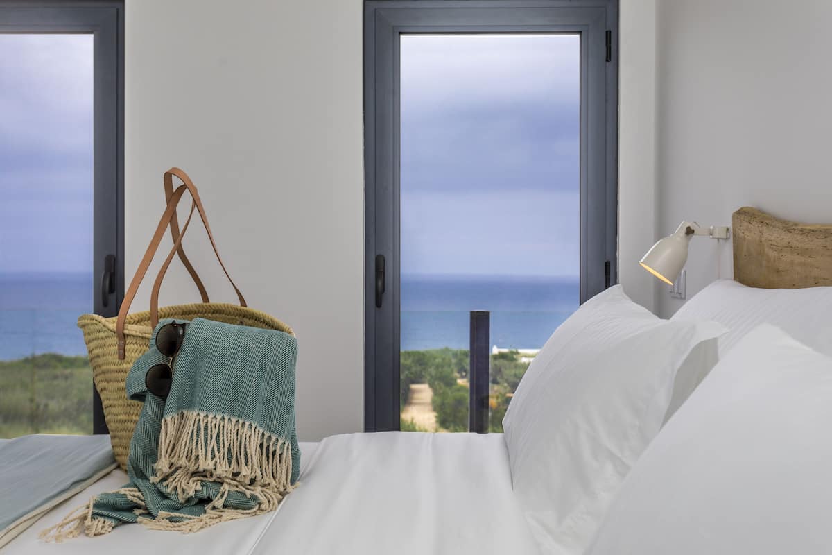 Quarto com cama, janela com vista para o mar, luminária e uma bolsa de tecido.