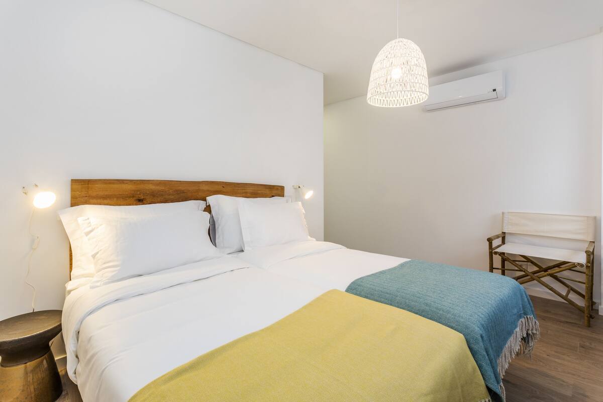 Uma cama com roupa de cama branca, cabeceira de madeira, luminária pendente e ar condicionado branco na parede.