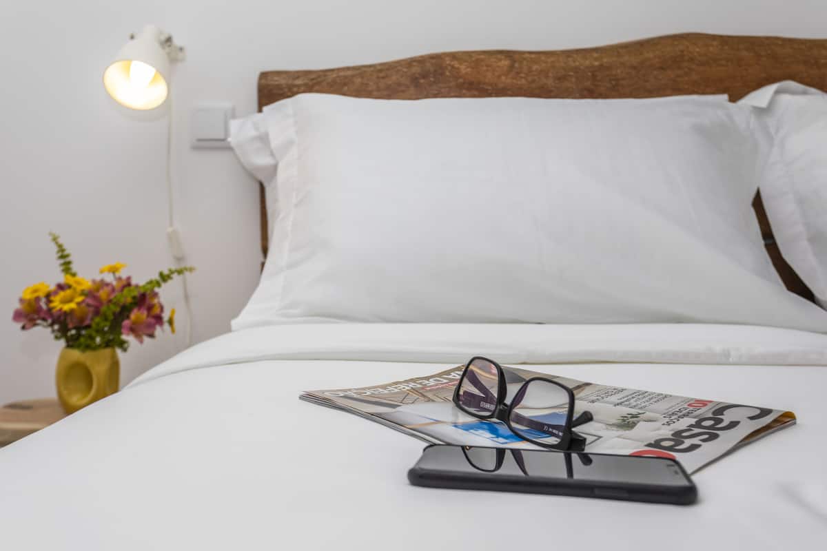 Cama arrumada com roupa de cama branca, um par de óculos sobre uma revista e um abajur.
