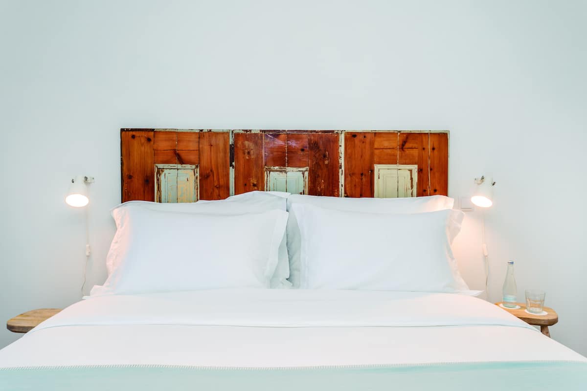 Cama com lençóis brancos, cabeceira de madeira, abajures e uma mesinha com copo e garrafa.