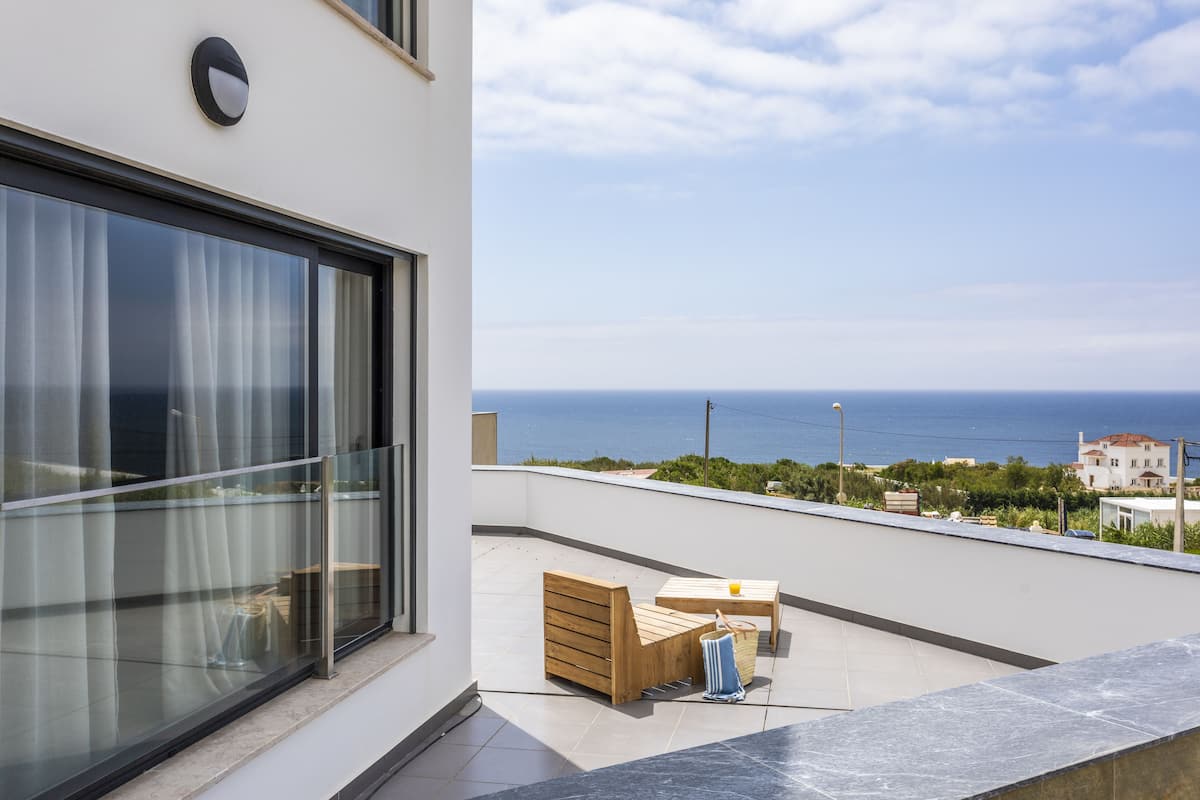 Uma varanda com um banco e mesa de madeira, porta de vidro e vista para o mar.