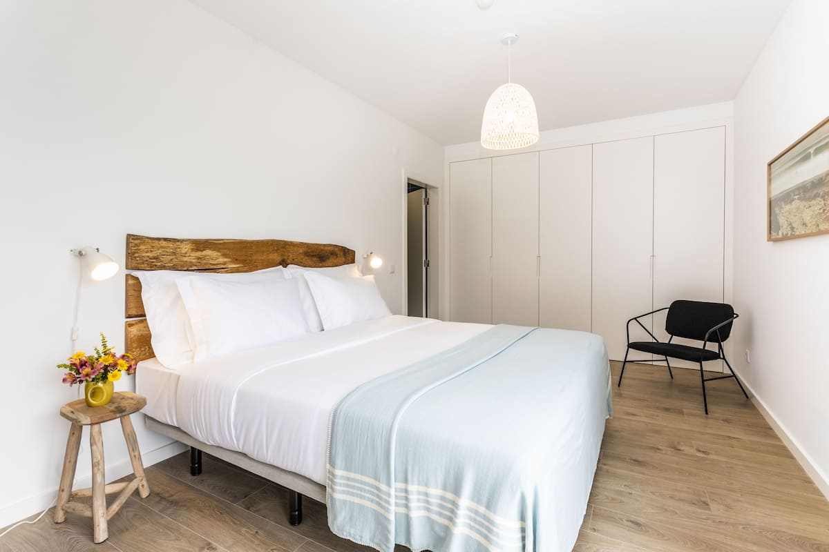 Quarto com cabeceira de madeira, roupa de cama branca, manta azul clara, um banquinho de madeira com um vaso de flores, uma cadeira preta e um guarda-roupa branco.