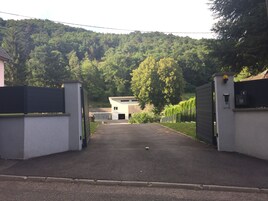 Uma entrada com portão leva a um edifício moderno com garagem, cercado por vegetação exuberante e colinas.