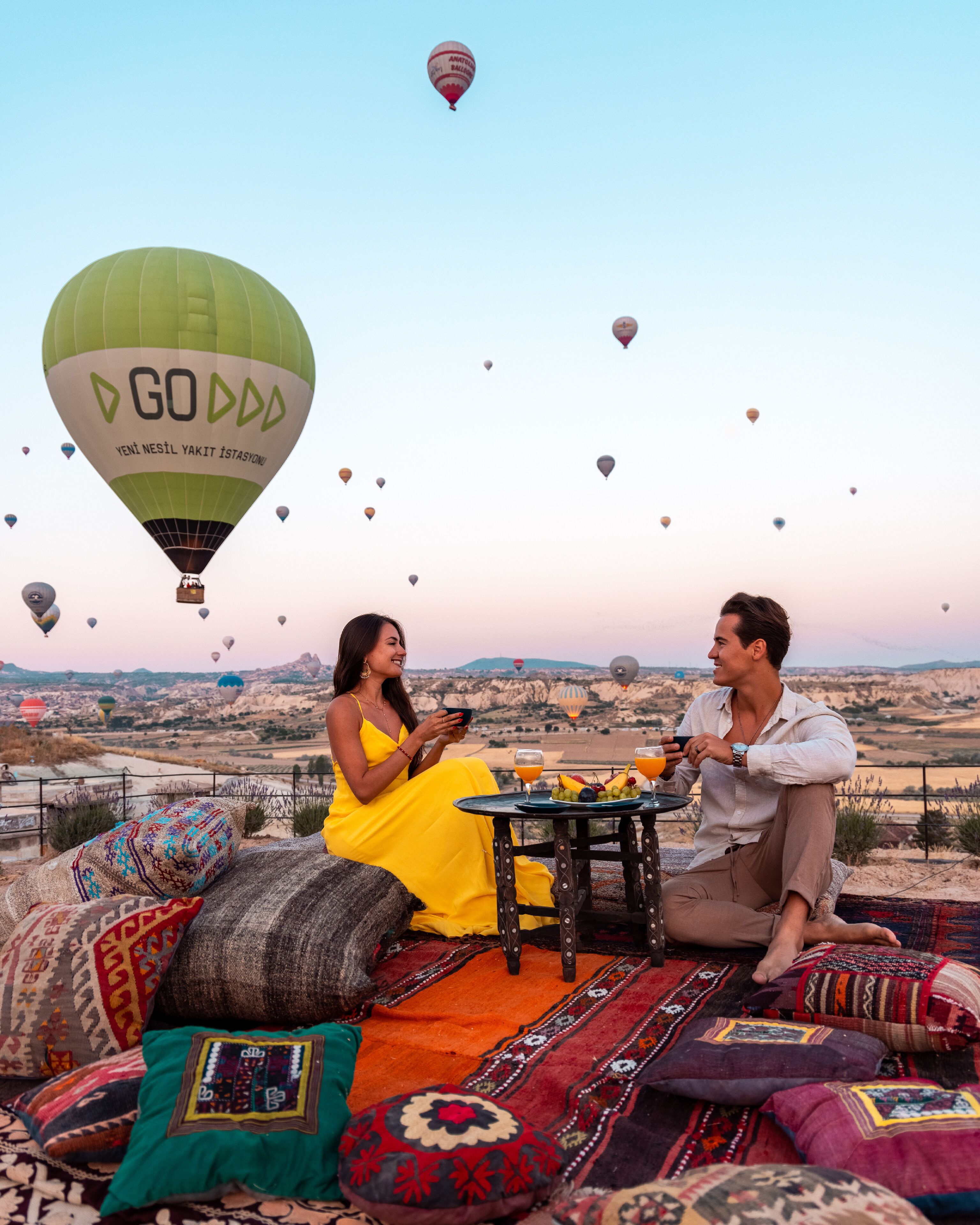 Um casal sentado num tapete colorido com uma mesa entre eles, rodeado por almofadas e uma vista panorâmica de um vale com balões de ar quente no céu.