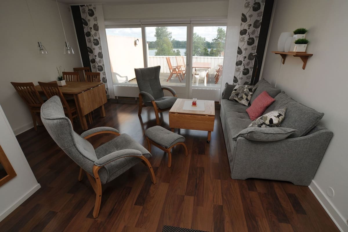 Uma sala de estar com um sofá cinzento, duas poltronas cinzas, uma mesa de centro de madeira, uma mesa de jantar com cadeiras e uma varanda com vista para árvores e água.