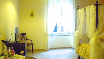 Quarto com cama, escrivaninha com computador, cadeira, janela com cortinas e vista para edifícios lá fora.