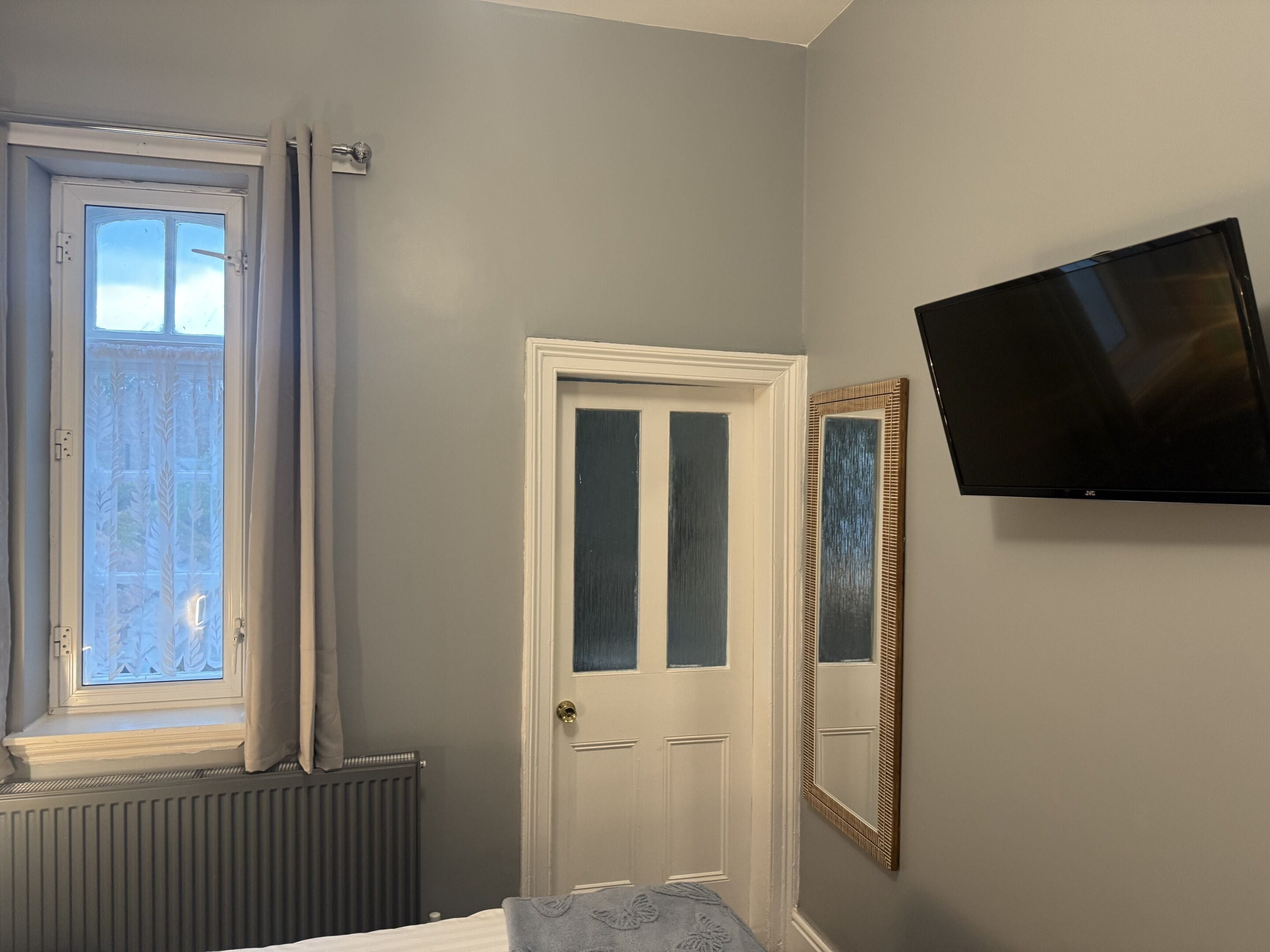 Quarto com porta branca, janela com cortinas, radiador e televisão na parede.