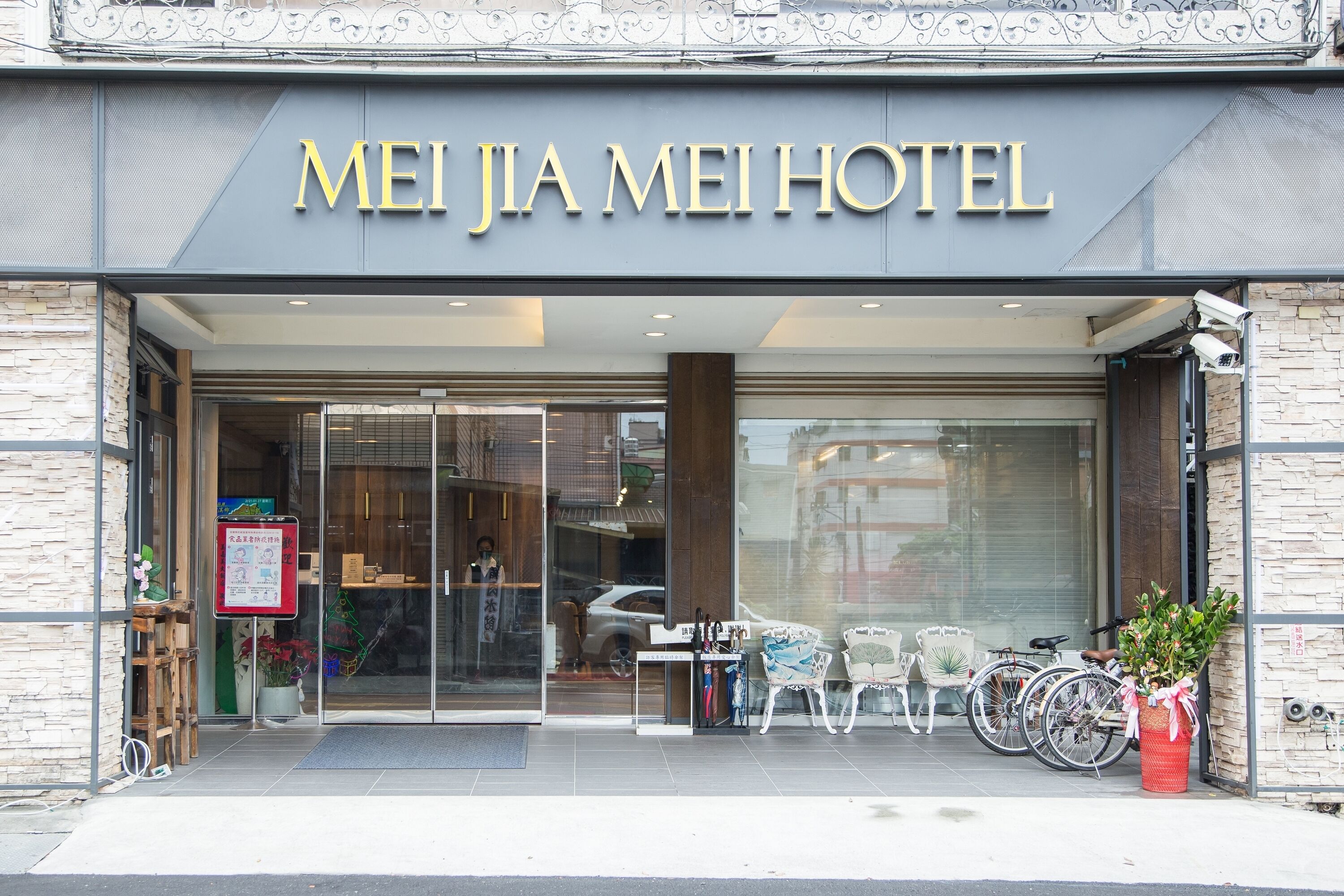 Entrada do Hotel Mei Jia Mei com fachada de vidro, uma placa e uma fileira de bicicletas na frente.