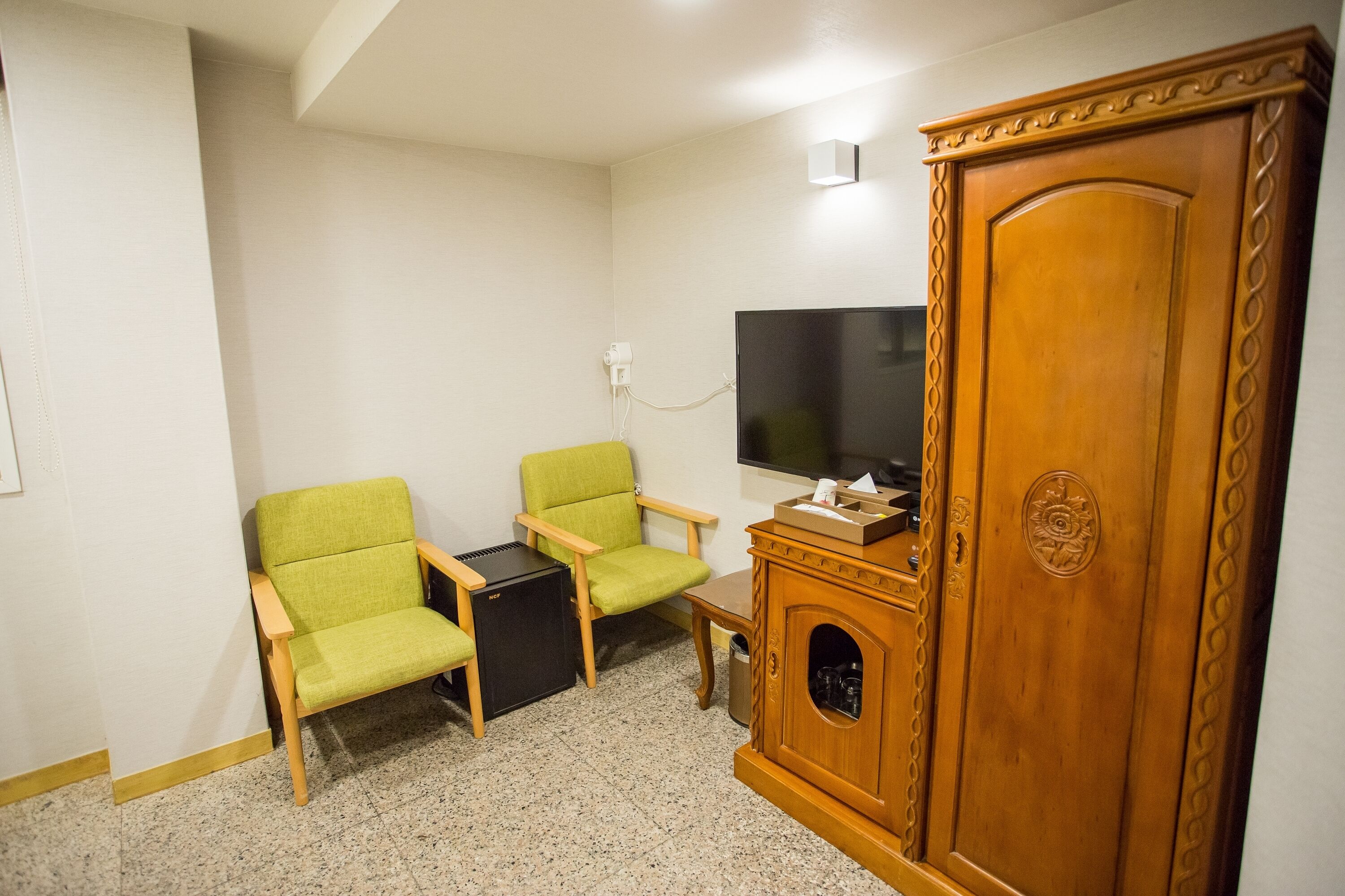 Quarto de hotel com duas poltronas verdes, um frigorífico preto pequeno, um móvel de madeira com televisão e chão em azulejos.