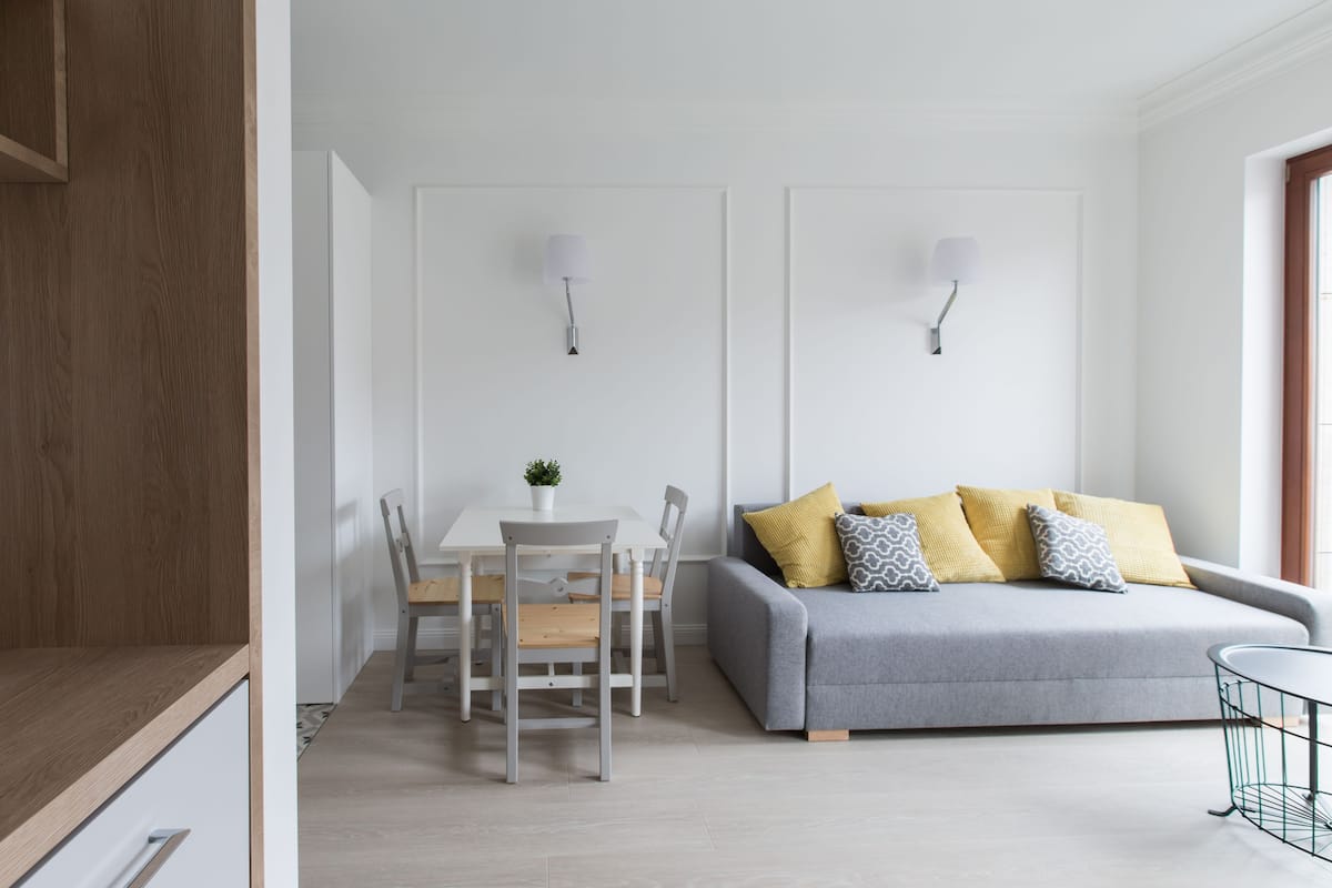 Uma sala de estar moderna com um sofá cinza, mesa de jantar de madeira e cadeiras. Há luminárias fixadas na parede e um pequeno vaso com planta sobre a mesa.