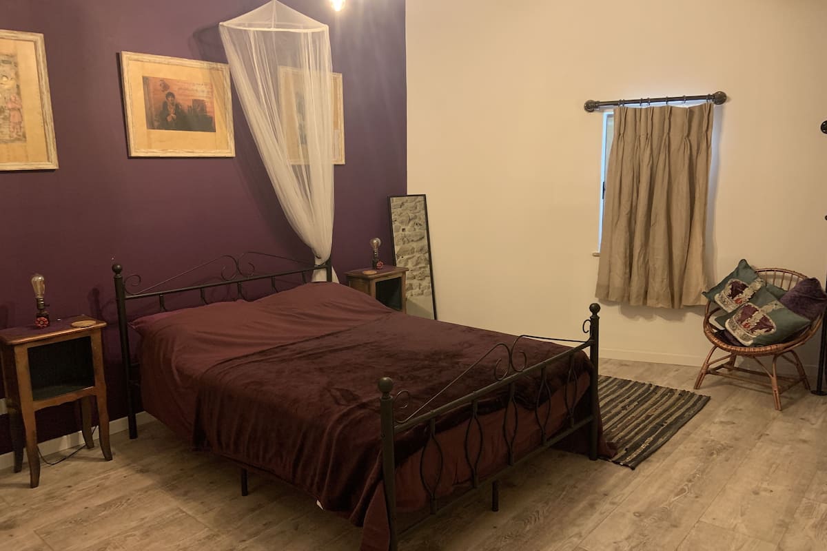 Um quarto com cama de dossel, mesa de cabeceira com abajur, cadeira de vime e janela com cortinas.