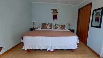 Quarto com cama, duas mesas de cabeceira, um criado-mudo, porta de madeira e um objeto decorativo fixado na parede.