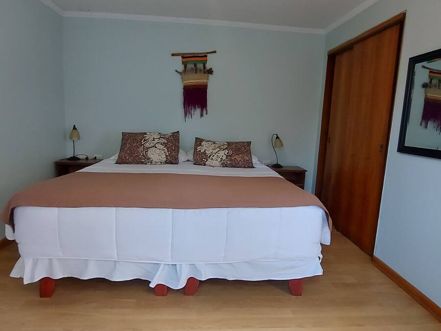 Quarto com cama, duas mesas de cabeceira, um criado-mudo, porta de madeira e um objeto decorativo fixado na parede.