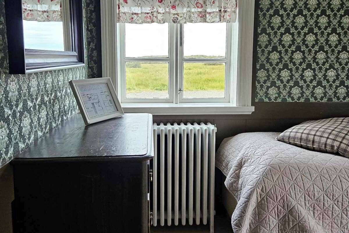Quarto com cama, cómoda, radiador e janela com cortinas florais.