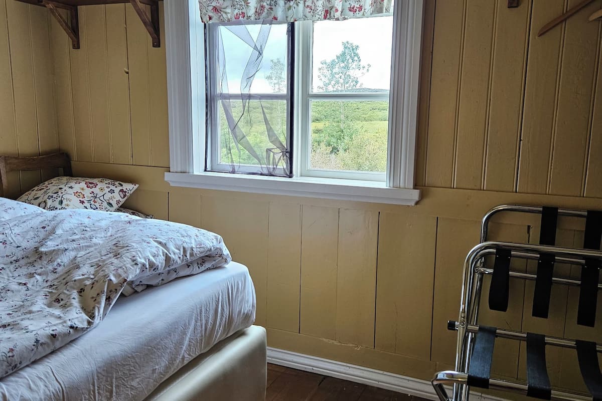 Quarto com piso de madeira, cama com roupa de cama floral, janela com cortinas florais e um suporte de metal.