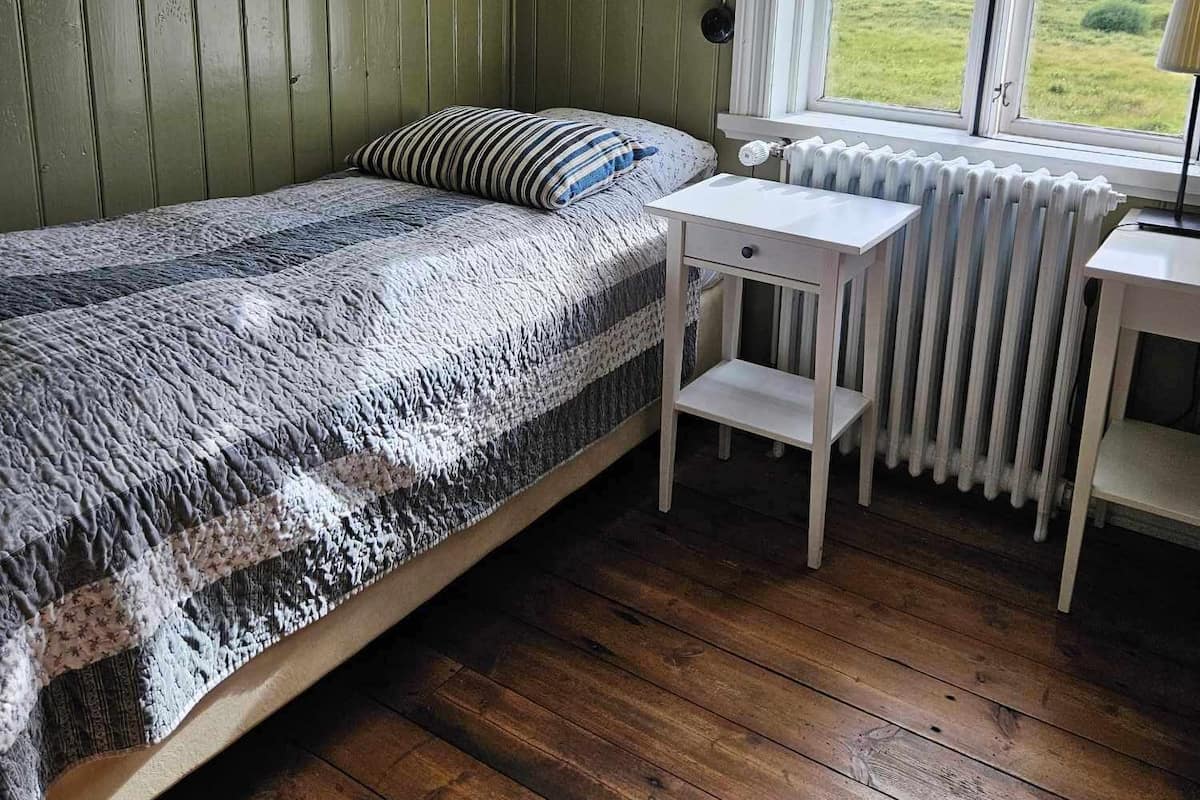 Quarto com uma cama, uma mesinha branca e janela com vista para uma paisagem verde.