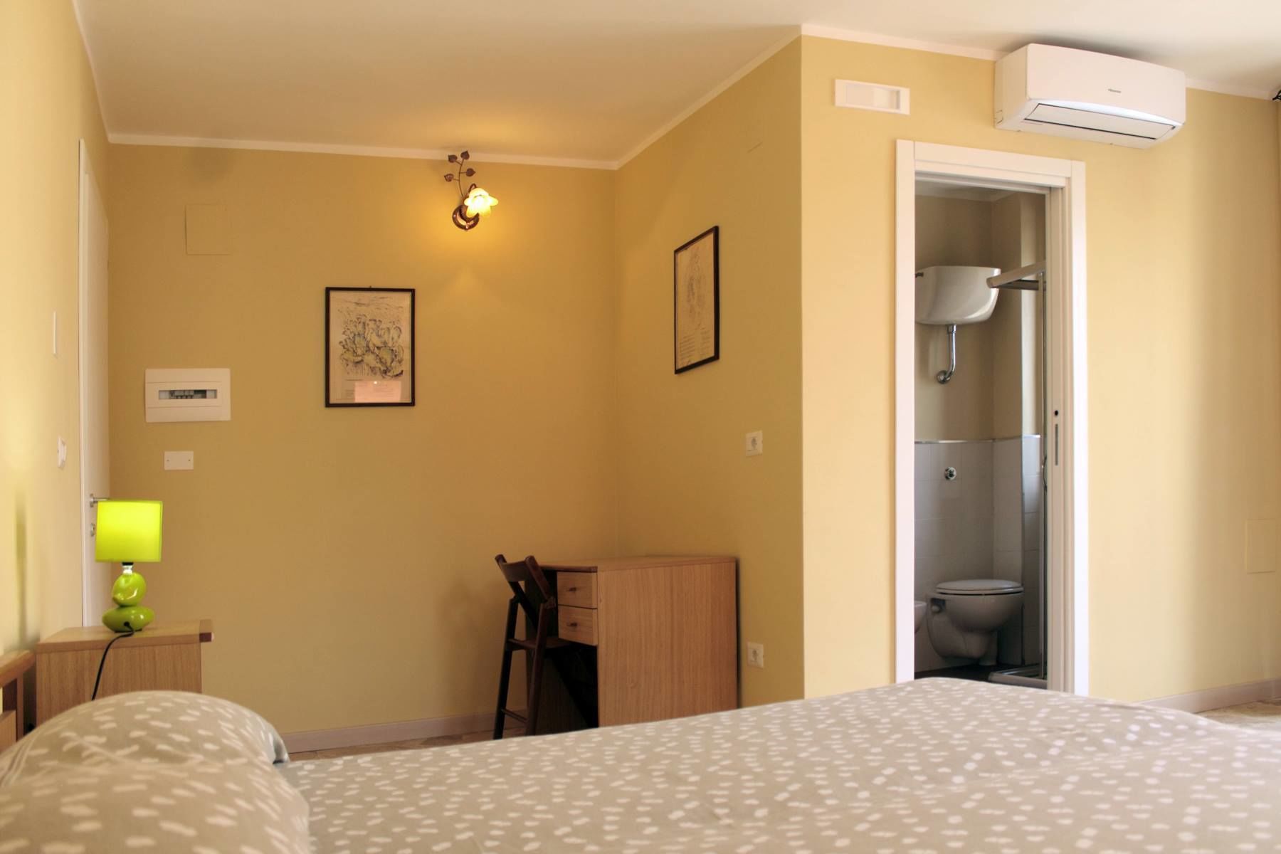 Quarto de hotel com cama, escrivaninha, cadeira, armário pequeno, luminária na parede, quadro emoldurado, vaso sanitário e unidade de ar condicionado.