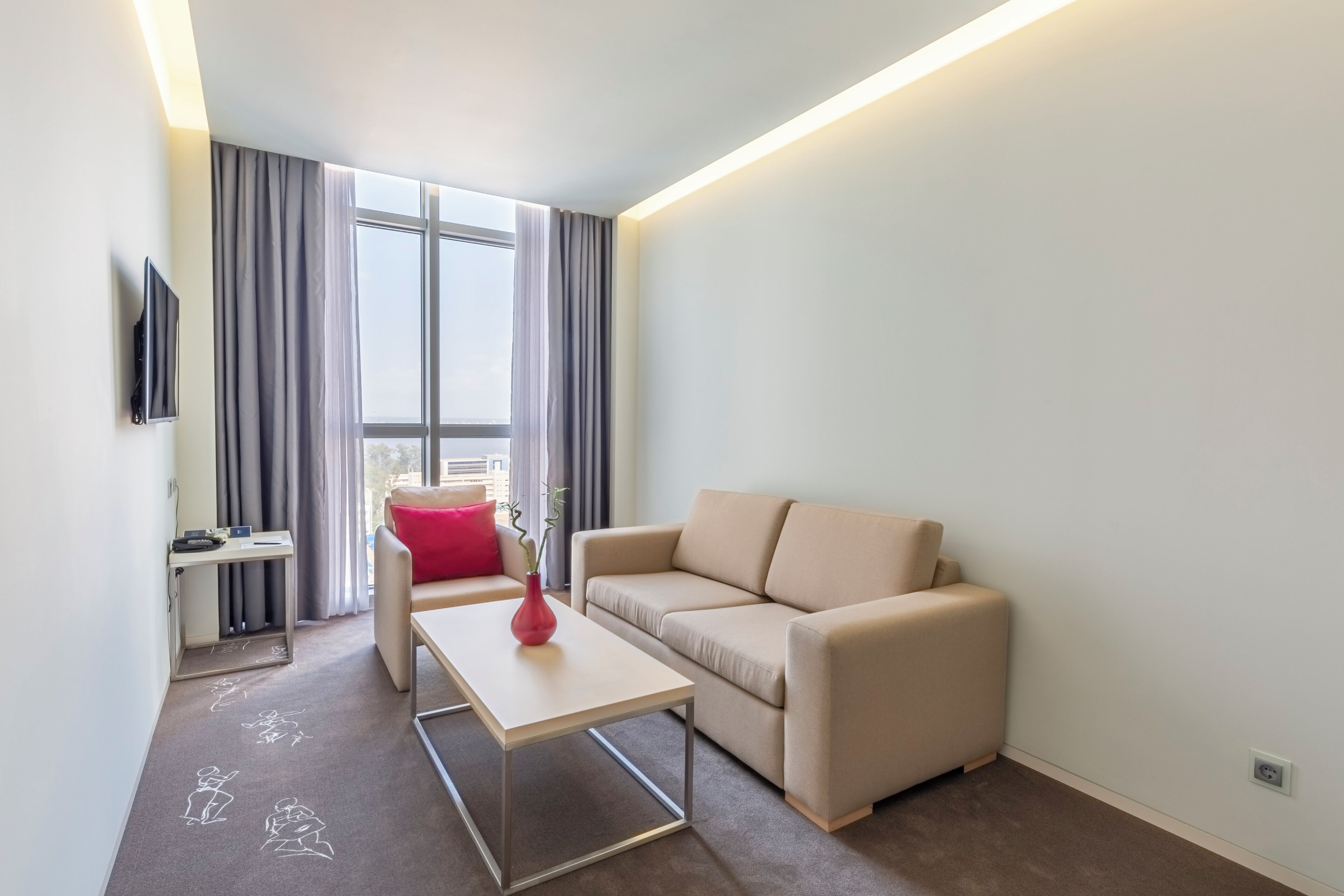 Quarto de hotel moderno com sofá, uma mesinha, televisão na parede e janela com cortinas.
