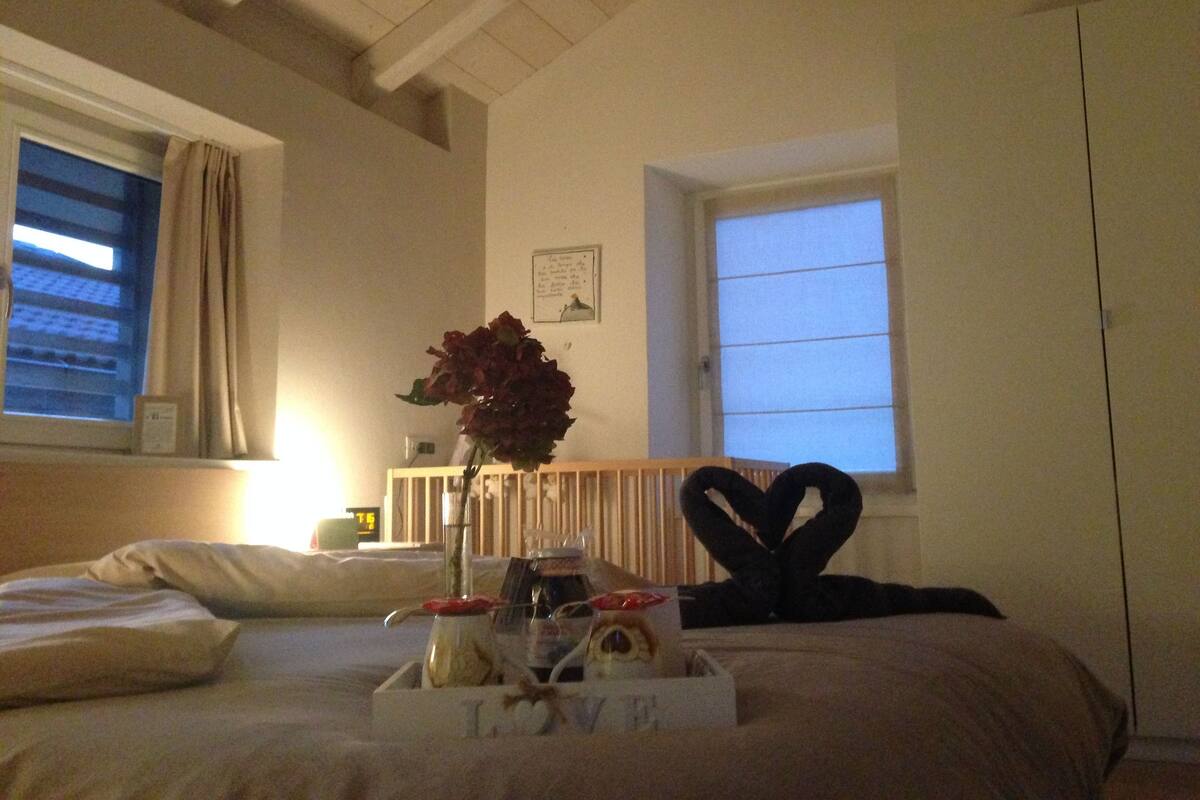 Quarto de hotel com cama, janela com cortinas, mesa de cabeceira com um vaso de flores e uma decoração em formato de coração na parede.