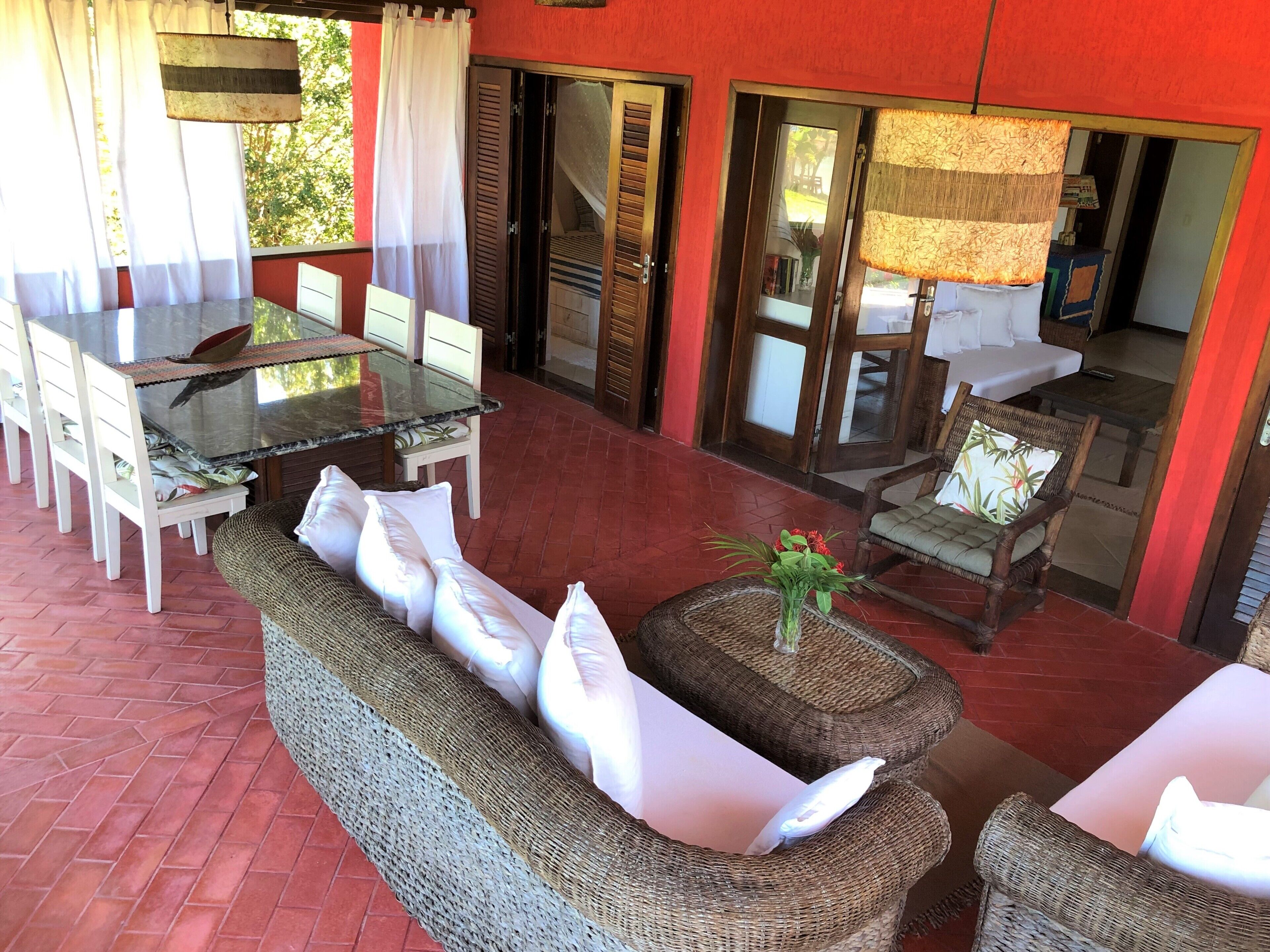 Uma área de estar coberta ao ar livre, com mesa de vidro, sofá e cadeiras de vime, integrada a um espaço de estar interno com sofá e abajur.