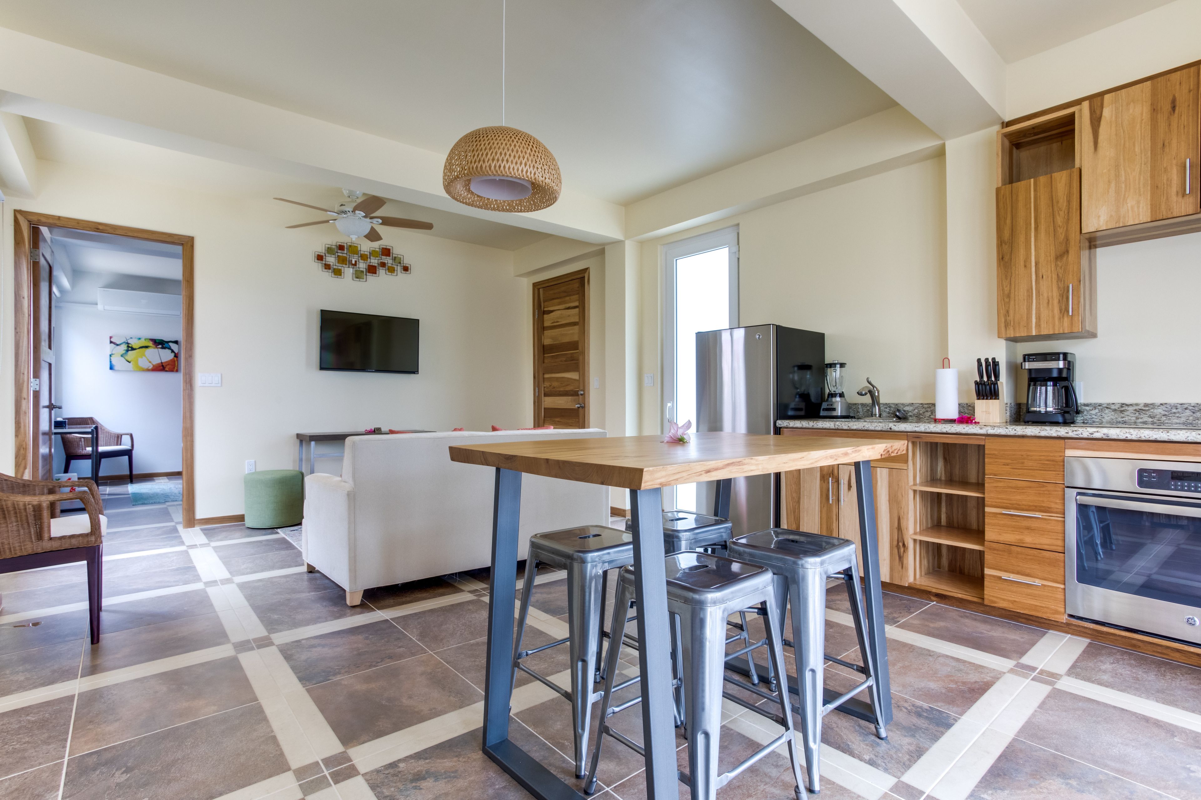 Cozinha moderna com mesa de madeira, banquetas, geladeira, cafeteira e televisão.