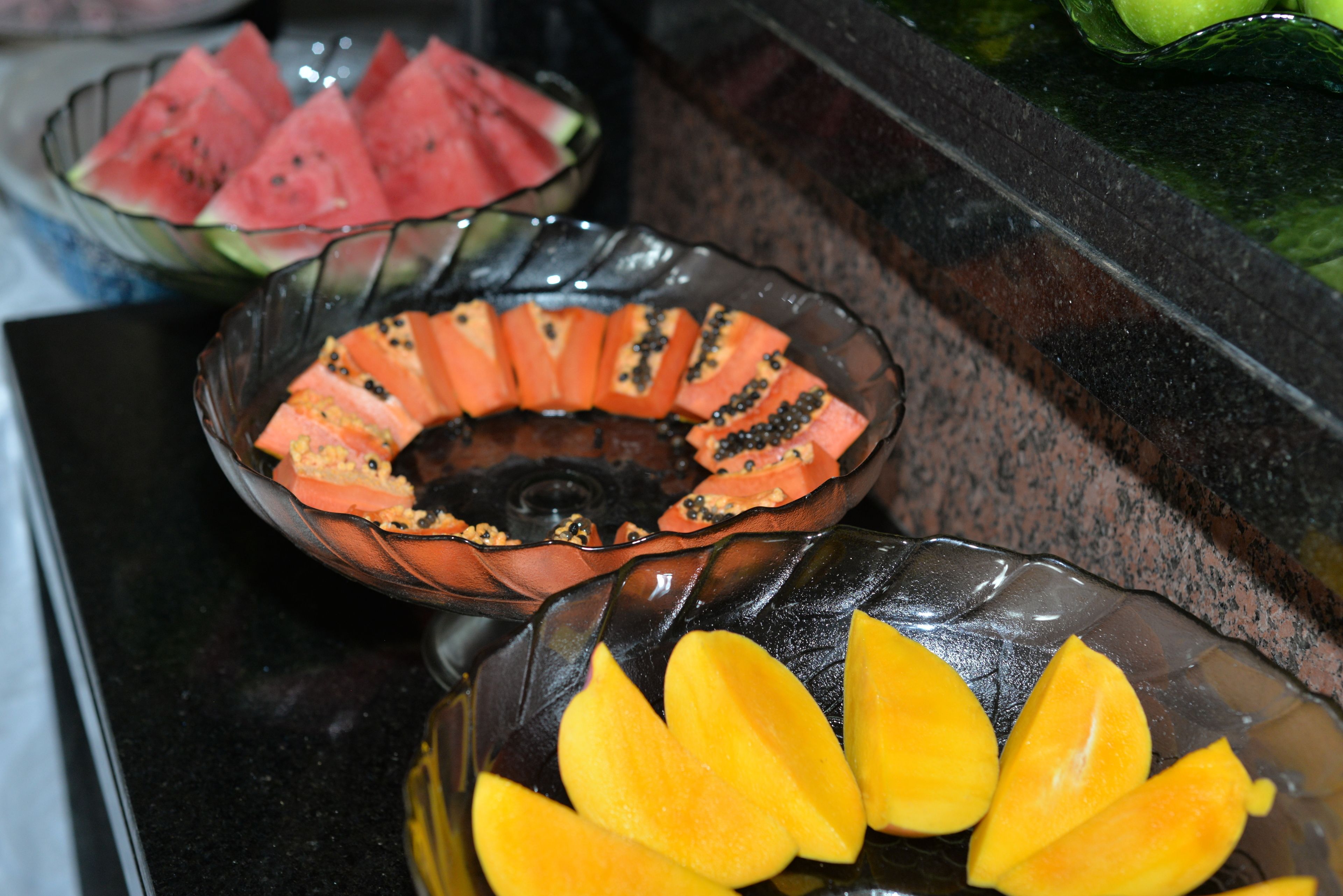 Uma tigela de vidro com frutas fatiadas, incluindo mamão e manga, sobre uma bancada de granito.