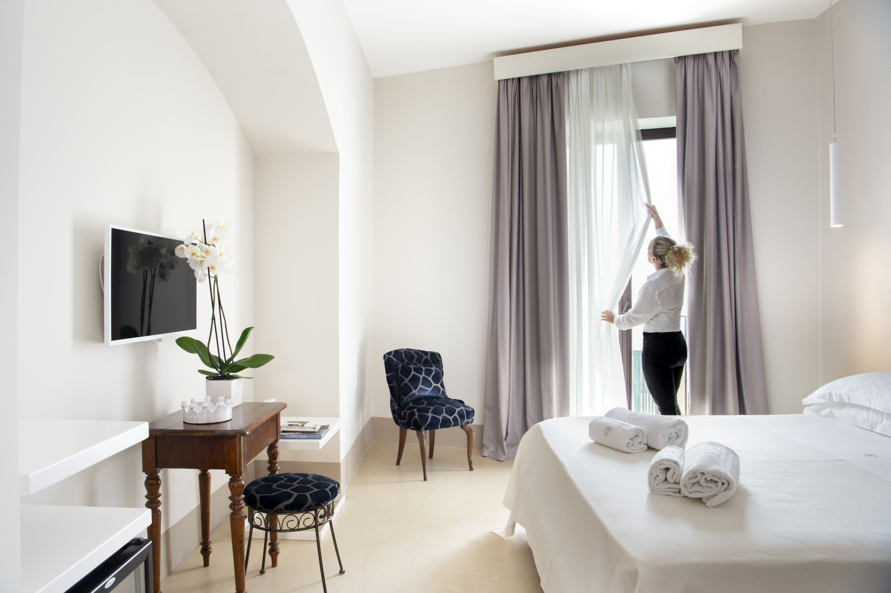 Quarto de hotel com uma cama, escrivaninha, cadeira, televisão e janela com cortinas.