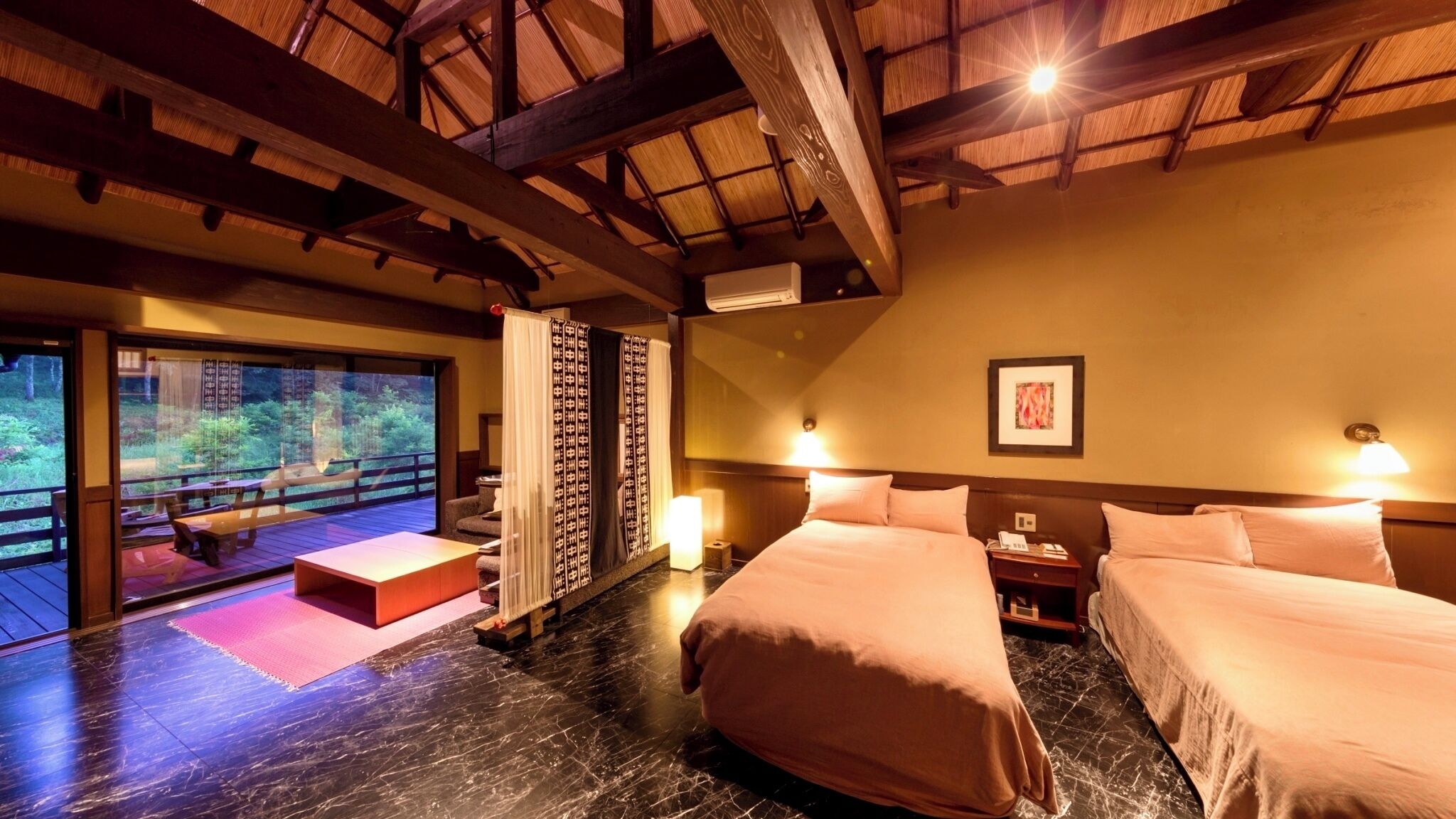 Quarto de hotel com uma cama grande, uma cama menor, uma escrivaninha, uma cadeira, uma janela com vista para um terraço e um teto com vigas de madeira.