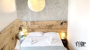 Quarto de hotel com cabeceira de madeira, cama com roupa de cama branca, duas toalhas enroladas, abajur e luminária na parede.