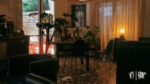 Uma sala de estar aconchegante com uma janela grande, uma mesa de jantar e várias cadeiras. Há plantas e um abajur sobre a mesa.