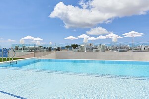 Piscina com guarda-sóis brancos e espreguiçadeiras, delimitada por um guarda-corpo de vidro e vista para o oceano.