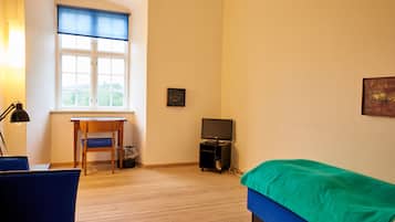Quarto com piso de madeira, uma escrivaninha com cadeira, televisão em um suporte e cama com manta verde.