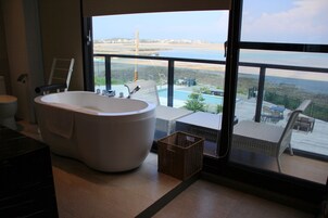 Casa de banho moderna com uma banheira grande, sanita e vista para a piscina e praia através de amplas janelas.