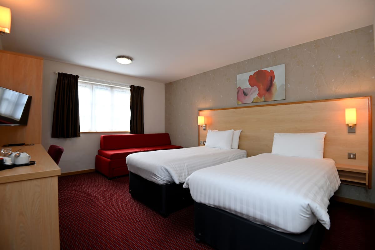 Quarto de hotel com duas camas, um sofá vermelho, uma escrivaninha com televisão e uma janela com cortinas.